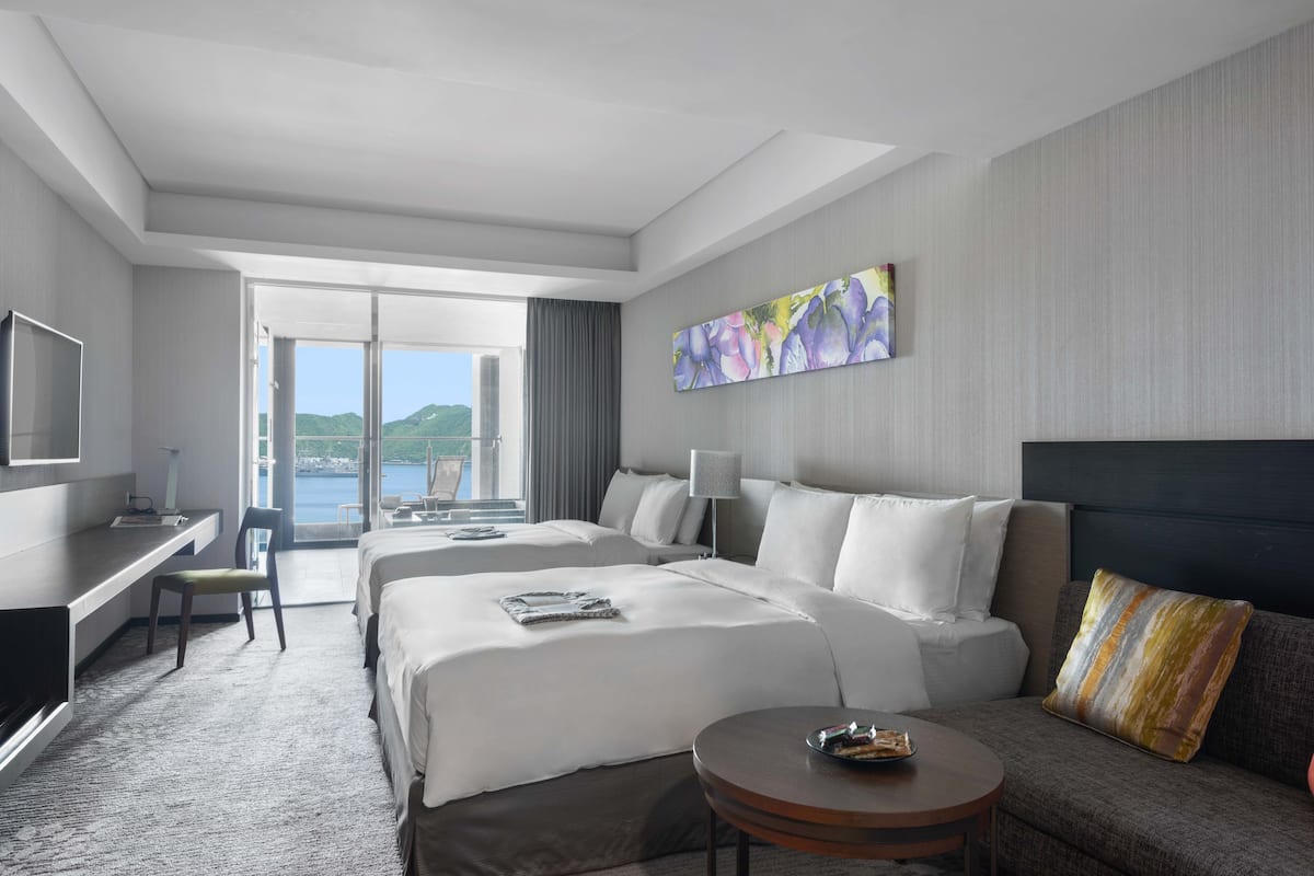 Quarto de hotel com duas camas, uma escrivaninha, uma cadeira, uma mesa redonda e uma janela grande com vista para o mar.