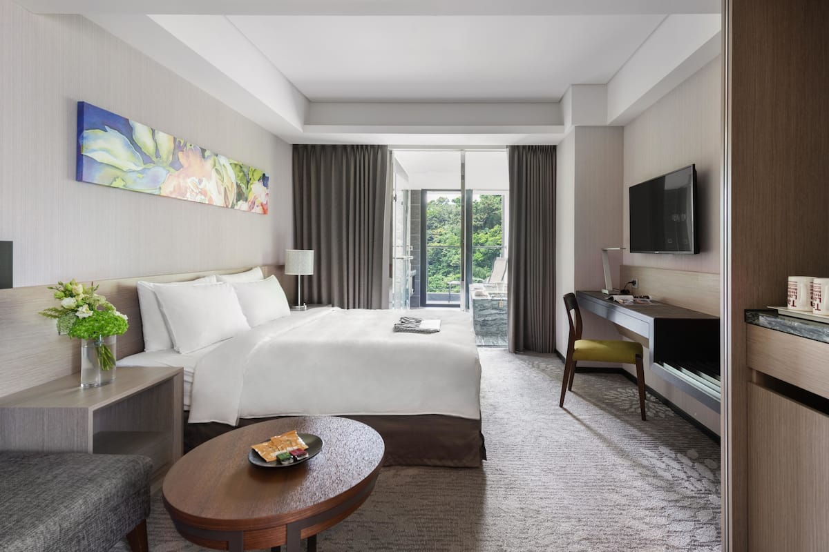 Quarto de hotel com cama, escrivaninha, televisão e vista para o verde lá fora.