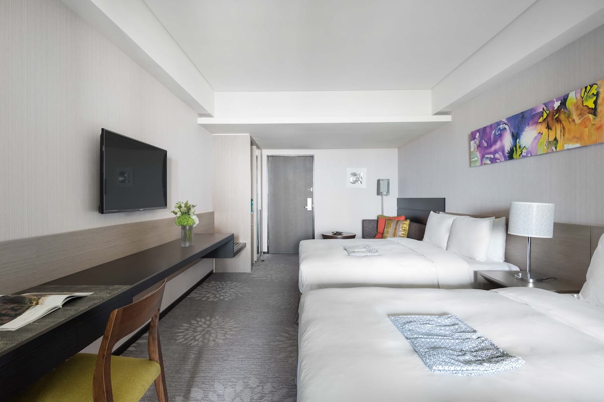 Quarto de hotel moderno com televisão de tela plana, escrivaninha com cadeira, duas camas, mesa de cabeceira, abajur e uma pintura abstrata colorida na parede.