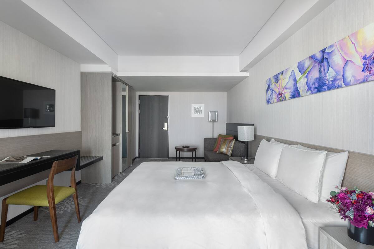 Quarto de hotel com cama, escrivaninha com cadeira, televisão, quadro na parede e mesa de cabeceira com luminária e flores.