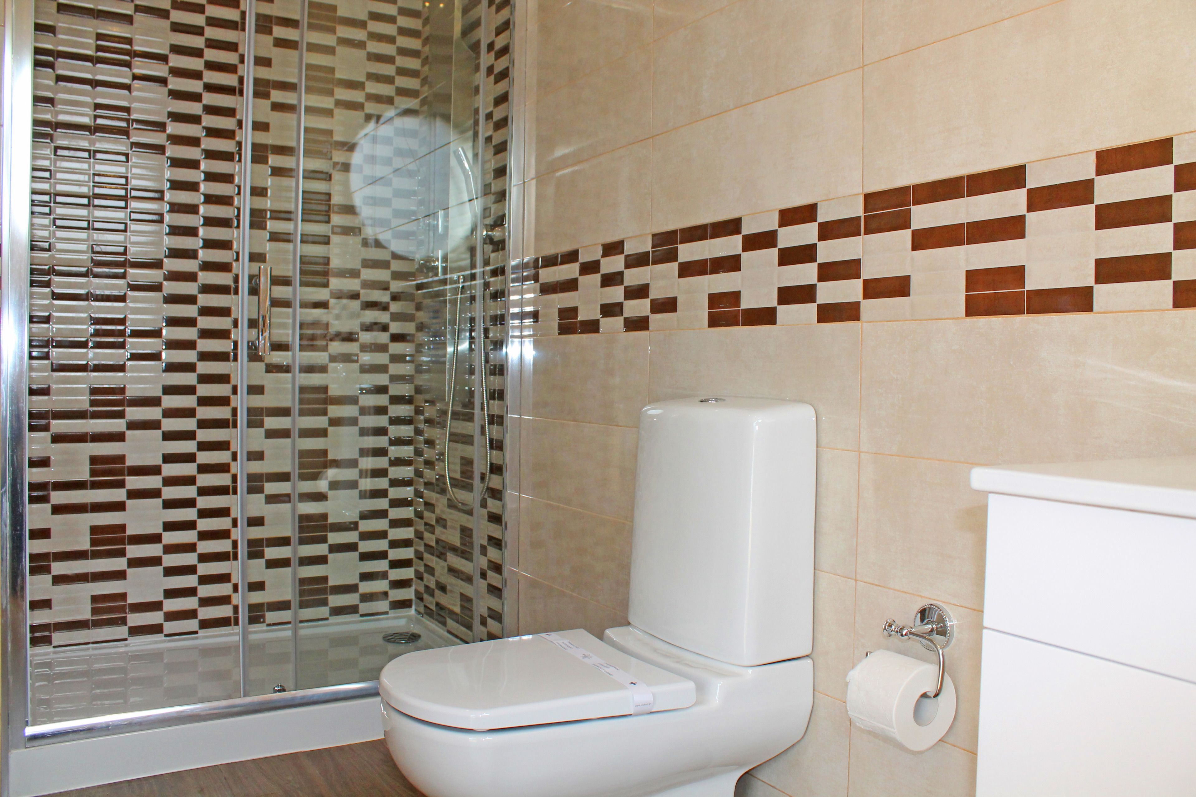 Casa de banho com cabine de duche em vidro, sanita e parede revestida a mosaicos com padrão xadrez.