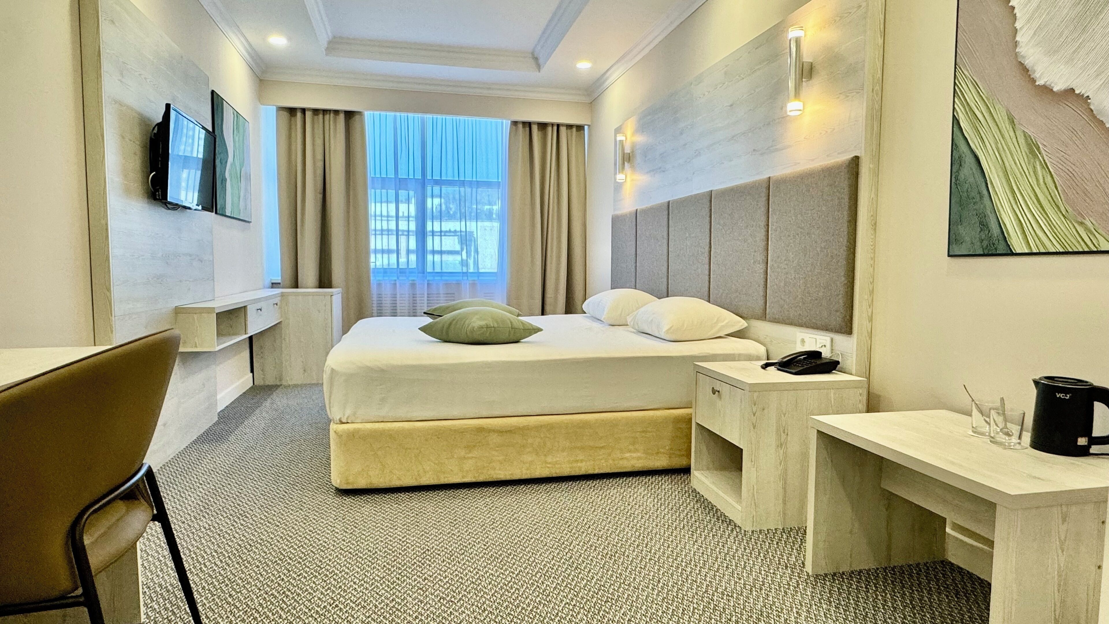 Quarto de hotel com uma cama, uma escrivaninha, uma cadeira, mesa de cabeceira com telefone e chaleira, e uma janela grande com cortinas.