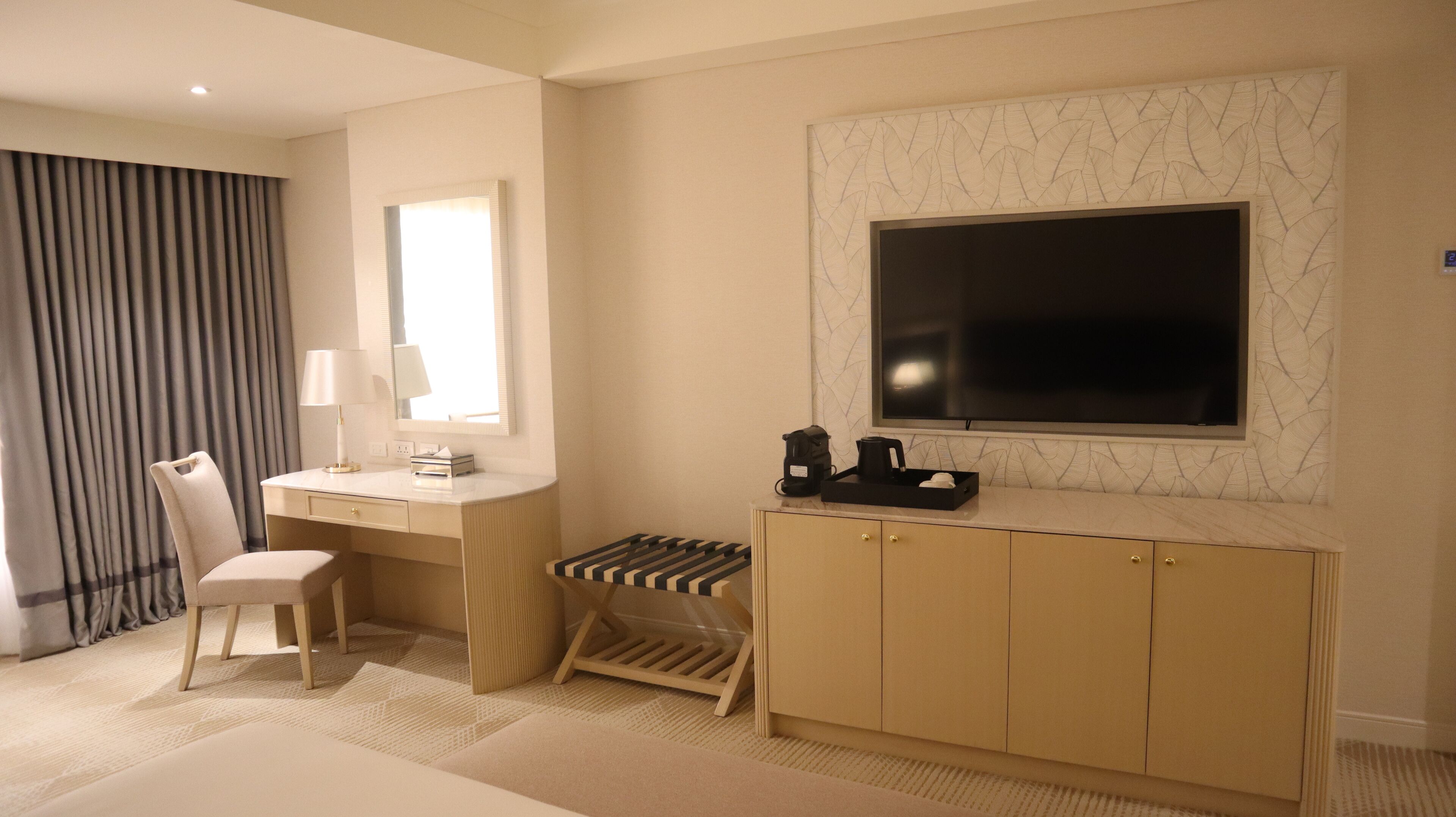 Quarto de hotel com escrivaninha, cadeira, televisão e um pequeno espaço para sentar.