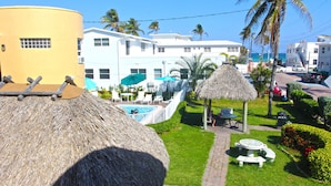 Uma área de piscina com estrutura de telhado de palha, um pequeno jardim com mesa e cadeiras, e vista para um edifício branco com várias janelas.