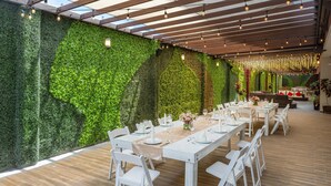 Uma sala de jantar com uma mesa comprida posta para seis pessoas, rodeada por cadeiras brancas, sob um teto de madeira com luminárias pendentes.