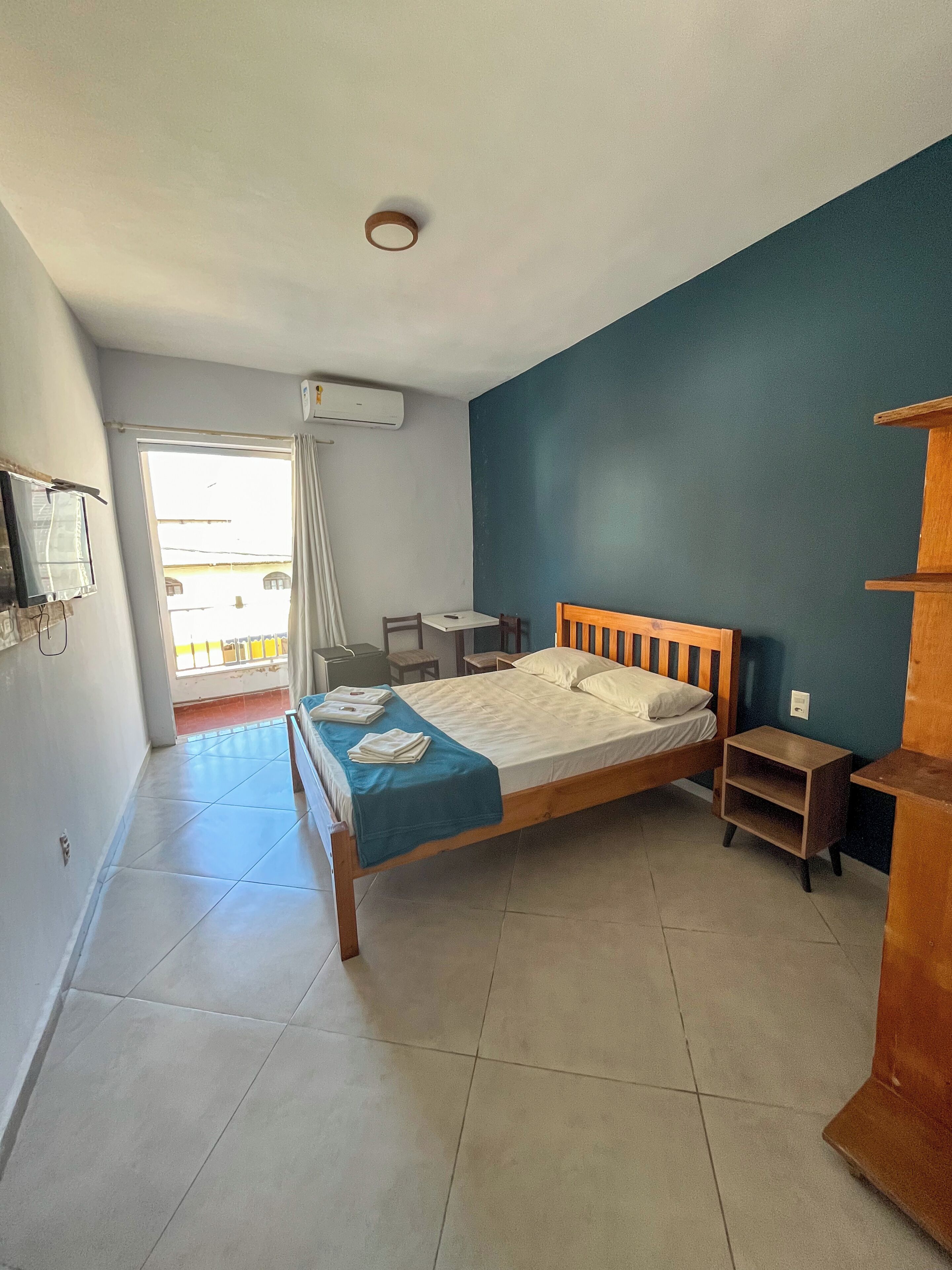 Quarto com cama, mesas de cabeceira, televisão e janela com cortinas.