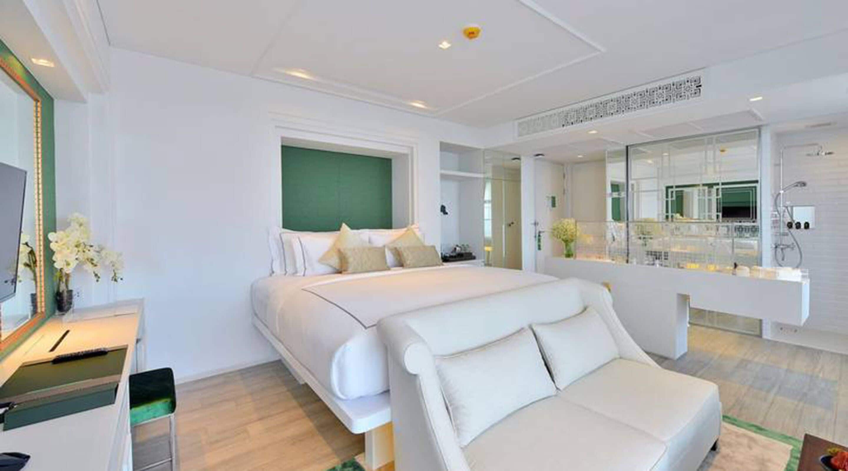 Quarto de hotel moderno com uma cama grande, um sofá, uma mesinha de vaidade com espelho e um banheiro visível através de um painel de vidro.