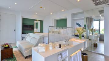 Casa de banho moderna com um espelho grande, lavatório e zona de duche. Existe um sofá branco na área de estar adjacente.