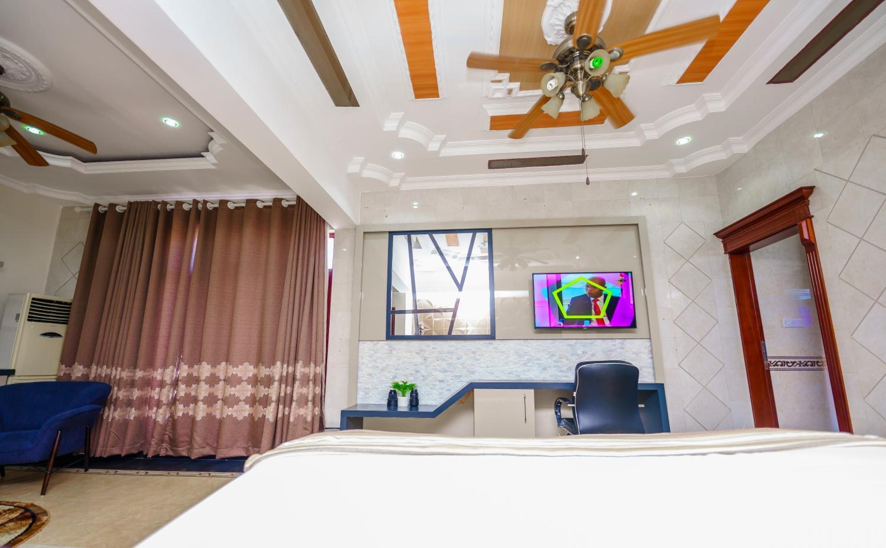 Quarto de hotel com cama, escrivaninha, cadeira, televisão e ventoinha no teto.