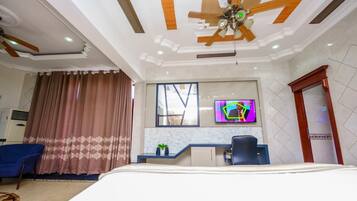 Quarto de hotel com cama, escrivaninha, cadeira, televisão e ventoinha no teto.