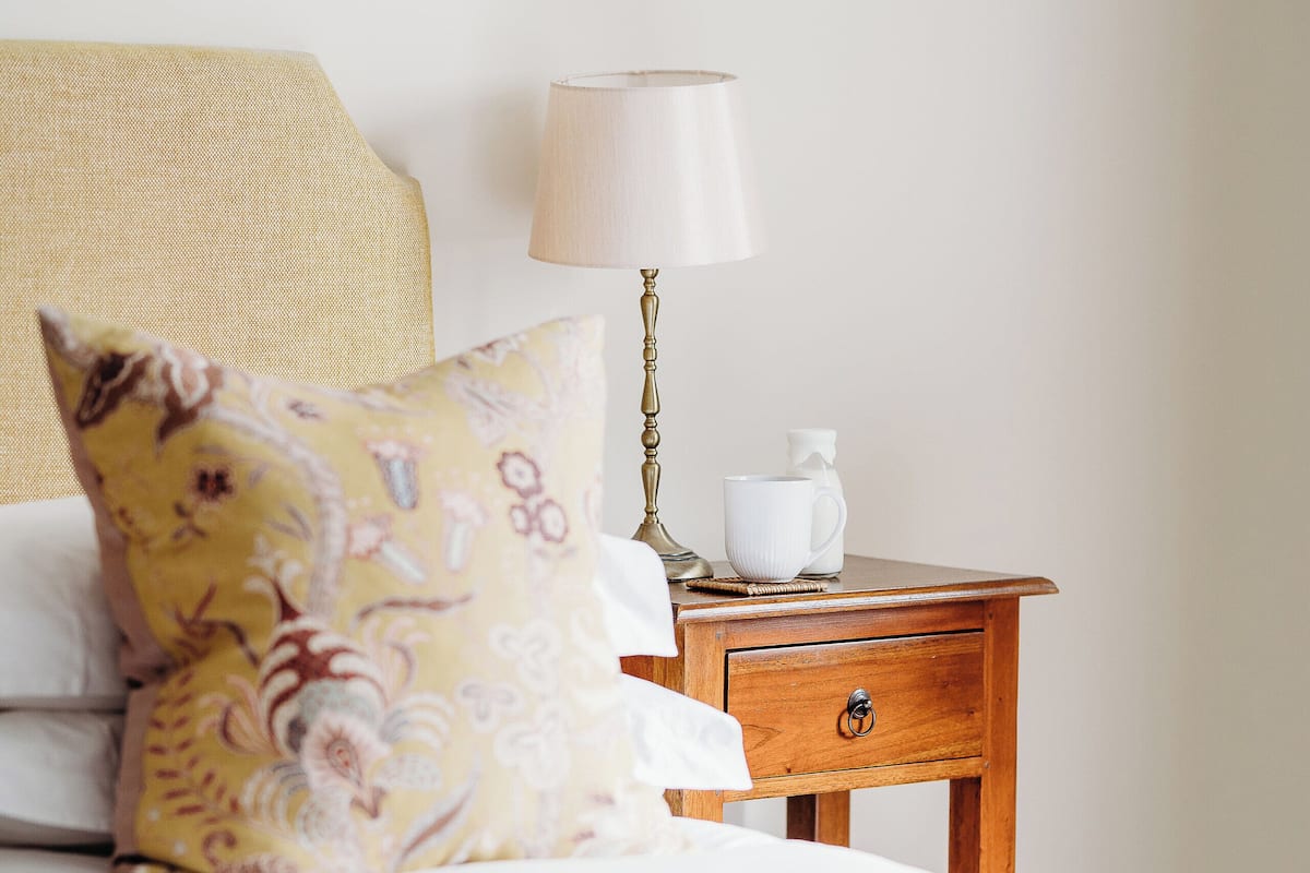 Uma cama bem arrumada com um travesseiro floral, uma mesa de cabeceira com abajur e uma chávena, e uma cómoda de madeira com uma gaveta.