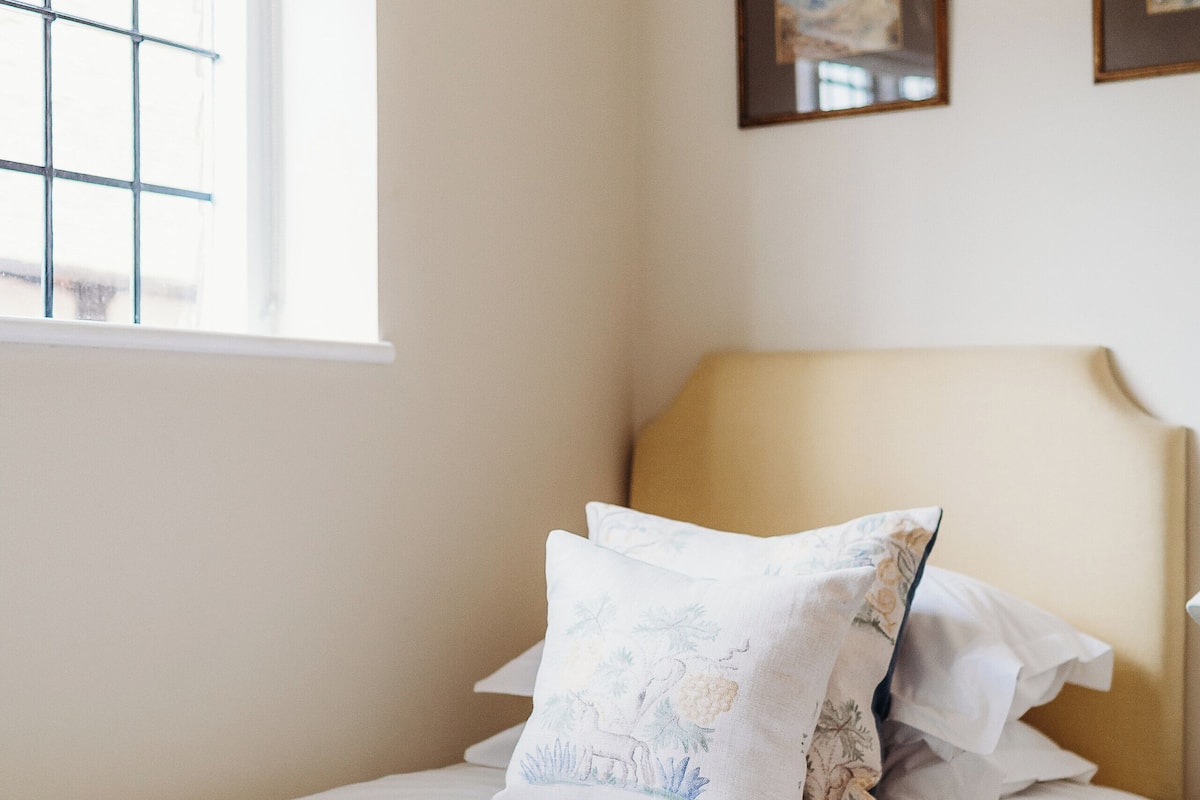 Cama bem arrumada com edredom floral branco e azul, cabeceira bege e janela com cortina branca.