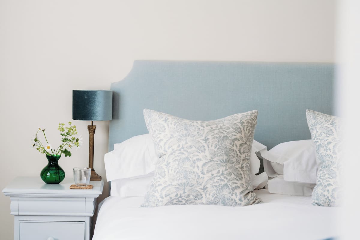 Cama bem arrumada com almofadas estampadas, mesa de cabeceira com um vaso de flores e um abajur.