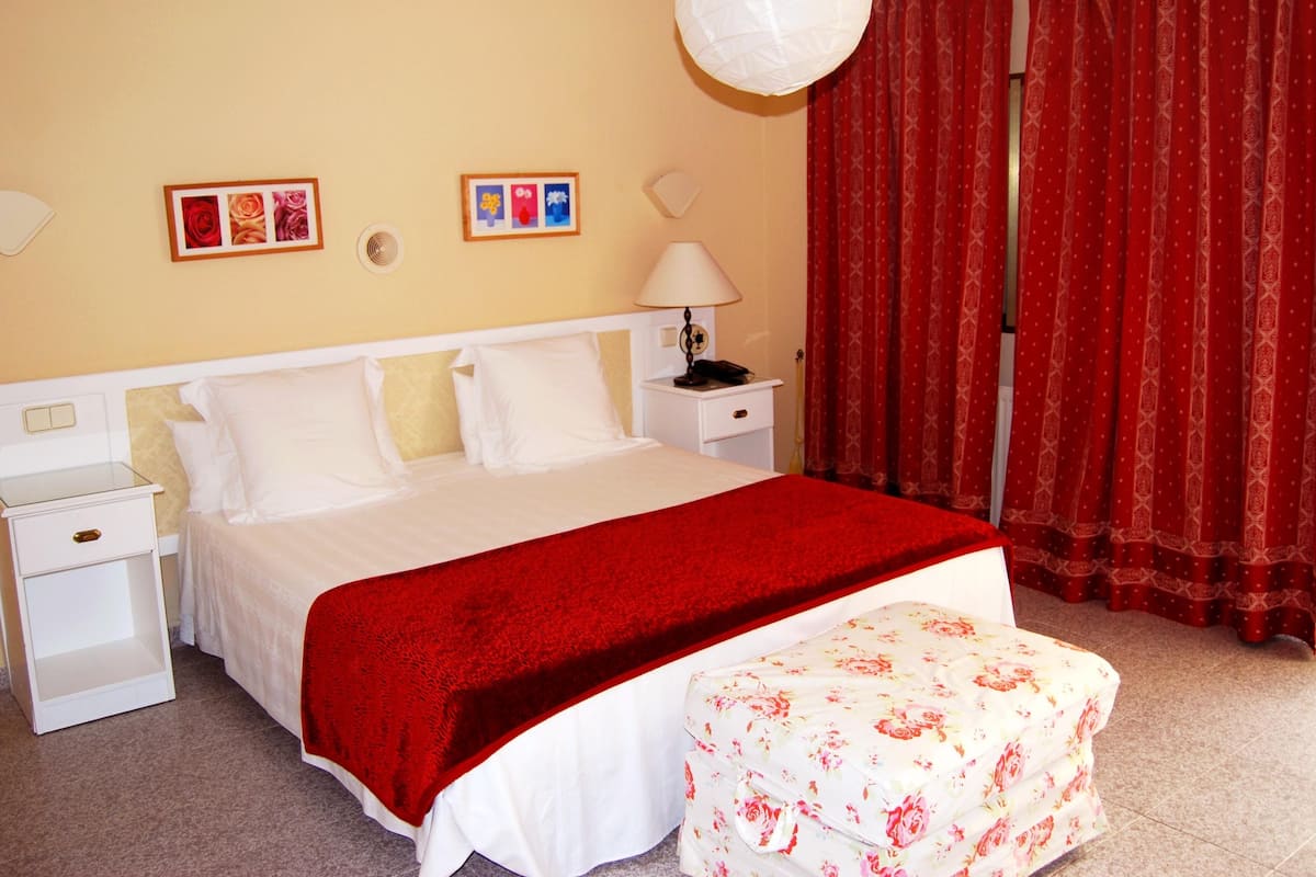 Quarto de hotel com cama, mesas de cabeceira, luminária, cortina vermelha e duas molduras na parede.