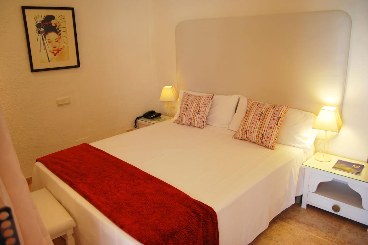 Cama bem arrumada com manta vermelha, duas luminárias de cabeceira e almofadas estampadas.