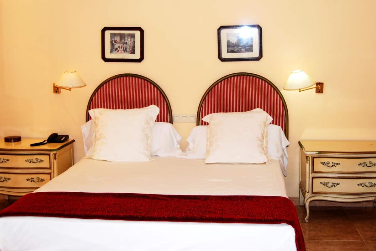Uma cama de casal com manta vermelha, duas mesas de cabeceira com abajures e duas molduras na parede.