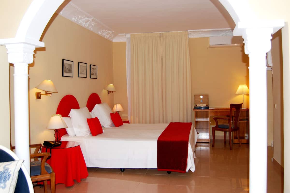 Quarto de hotel com uma cama, uma escrivaninha e uma cadeira. Há duas luminárias fixadas na parede e um telefone na escrivaninha.