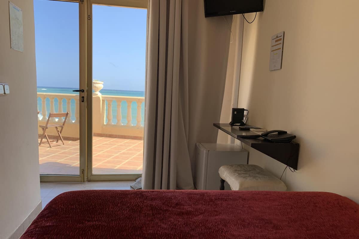 Quarto de hotel com cama, escrivaninha com telefone e vista para o mar através de uma porta de vidro.