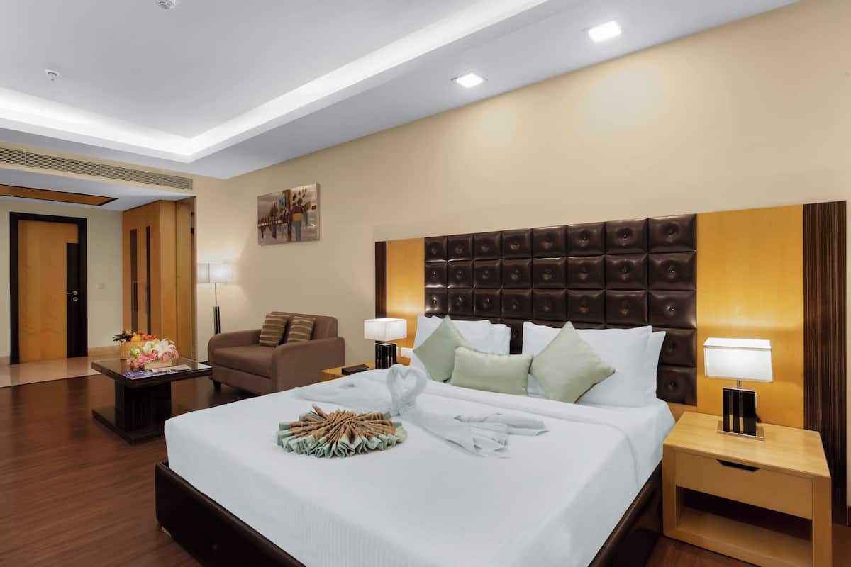 Quarto de hotel com uma cama grande, cabeceira marrom, sofá marrom, uma pequena mesa com abajur e um quadro na parede.