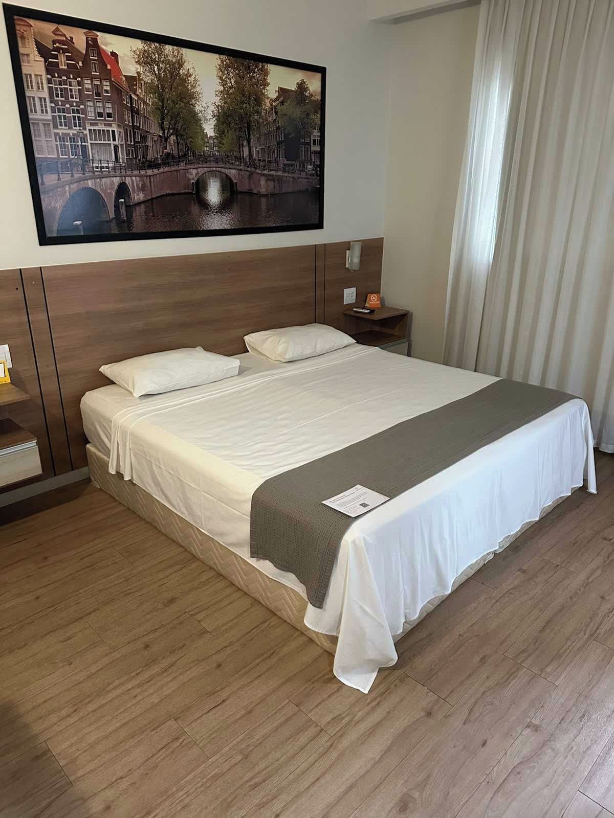 Quarto de hotel com cama grande, cabeceira de madeira, mesa de cabeceira, luminária na parede e pintura de uma paisagem urbana.