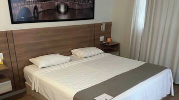 Quarto de hotel com cama grande, cabeceira de madeira, mesa de cabeceira, luminária na parede e pintura de uma paisagem urbana.