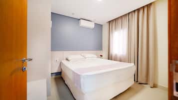 Quarto de hotel com uma cama, ar-condicionado instalado no teto, janela com cortinas e porta de madeira.