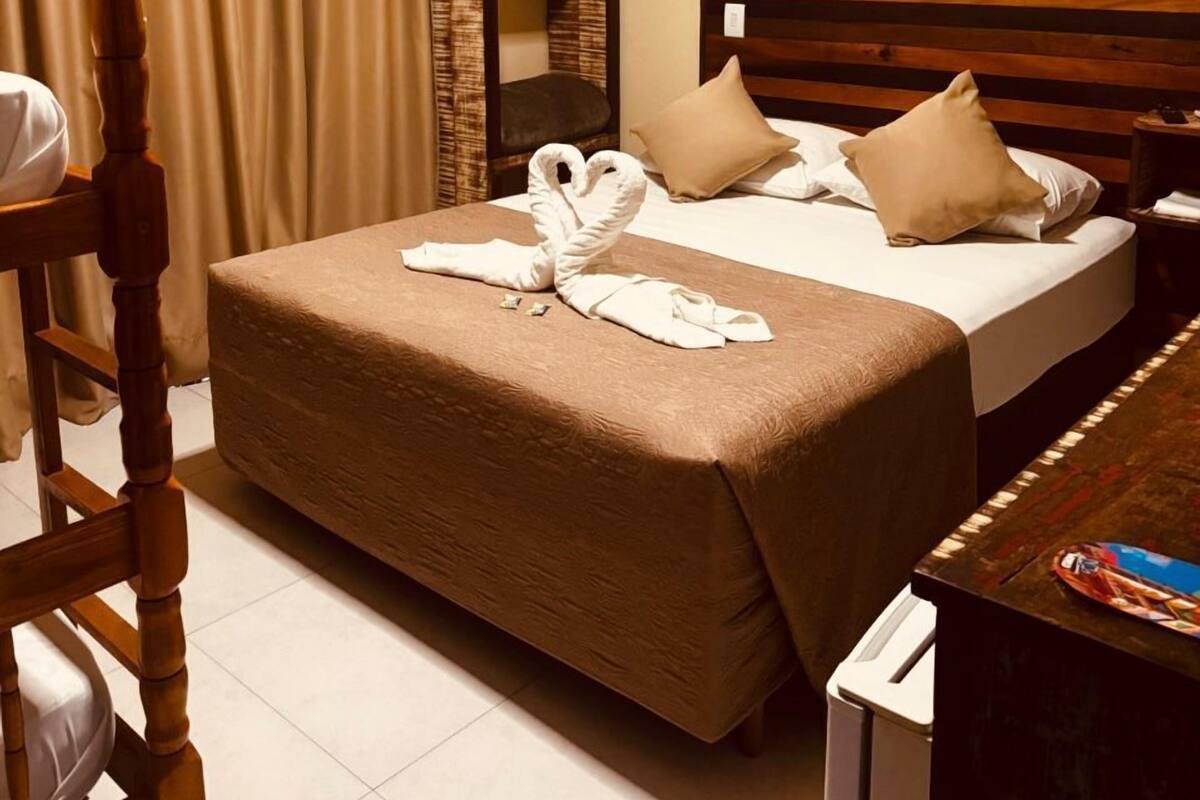 Quarto de hotel com uma cama grande, duas almofadas, cabeceira de madeira, mesa de cabeceira, espelho e uma cadeira de madeira.