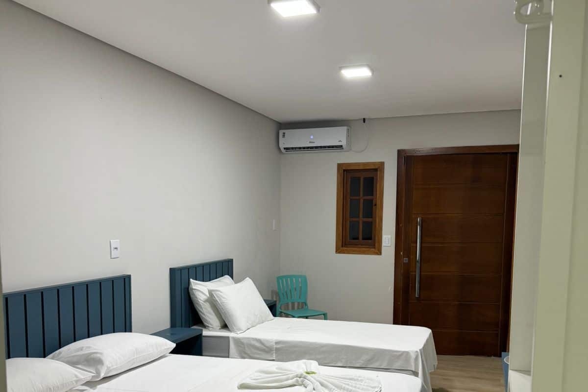 Quarto de hotel com duas camas, uma porta de madeira, uma janela e um aparelho de ar condicionado.