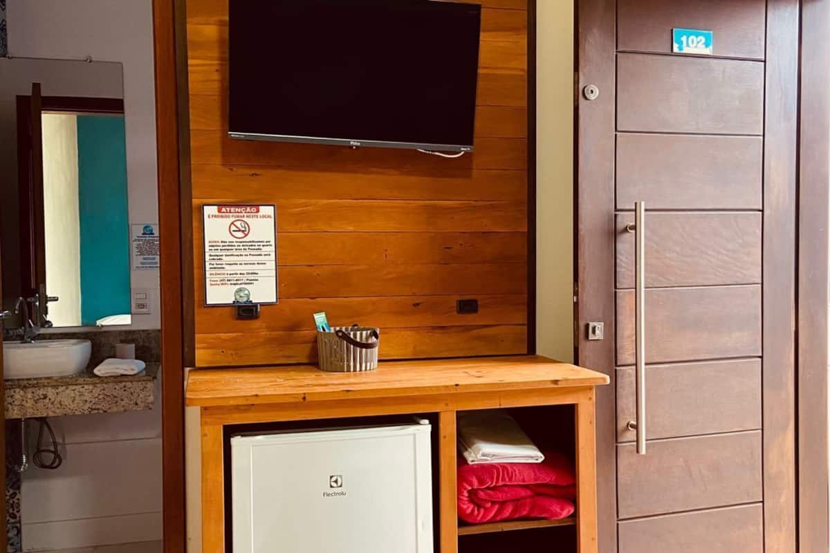Quarto de hotel com um armário de madeira, uma televisão montada acima, um mini frigorífico, uma porta com placa de número e um banheiro visível ao fundo.
