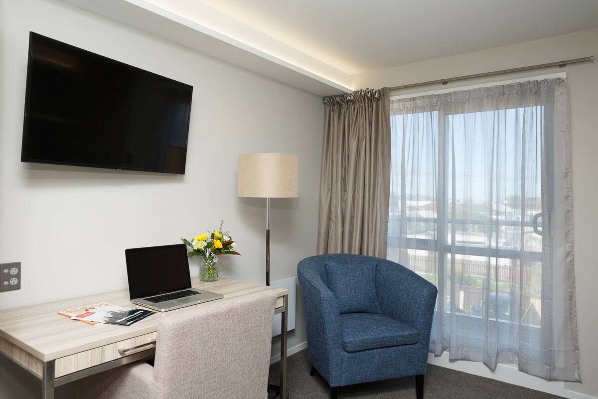 Quarto de hotel com televisão de tela plana na parede, uma escrivaninha com um laptop, uma cadeira, um abajur e uma janela com cortinas finas.