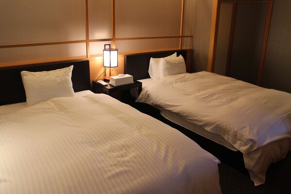 Quarto de hotel com duas camas, cada uma com lençóis e almofadas brancas. Há uma mesinha de cabeceira com abajur e caixa de lenços. O quarto possui cabeceira de madeira e um esquema de cores neutras.