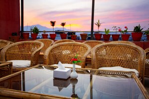 Uma sala de jantar com cadeiras de vime e uma mesa, um vaso com flores e vista para o pôr do sol através de grandes janelas.