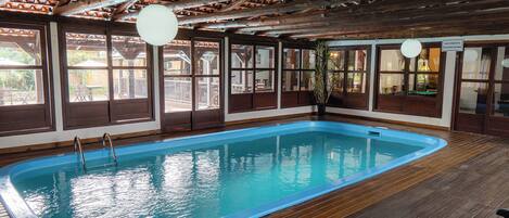 Piscina interna com deck de madeira, grandes janelas e teto com vigas aparentes.