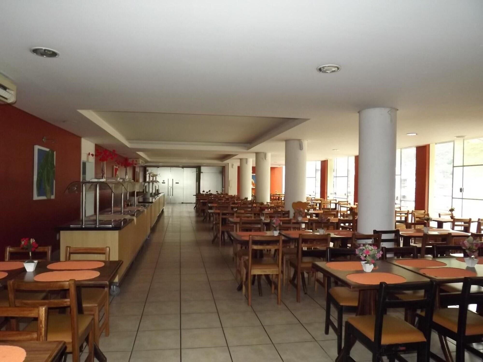 Uma sala de jantar ampla, com várias mesas e cadeiras, um balcão central de serviço e grandes janelas que proporcionam luz natural.