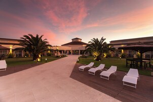 Pátio de hotel com cadeiras de lounge, palmeiras e um edifício com telhado em azulejos.