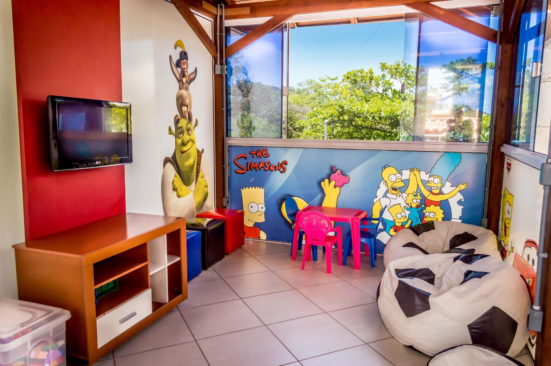 Uma sala de jogos infantil com televisão, móveis coloridos e painéis de parede com desenhos de 'The Simpsons'.