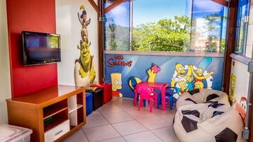 Uma sala de jogos infantil com televisão, móveis coloridos e painéis de parede com desenhos de 'The Simpsons'.