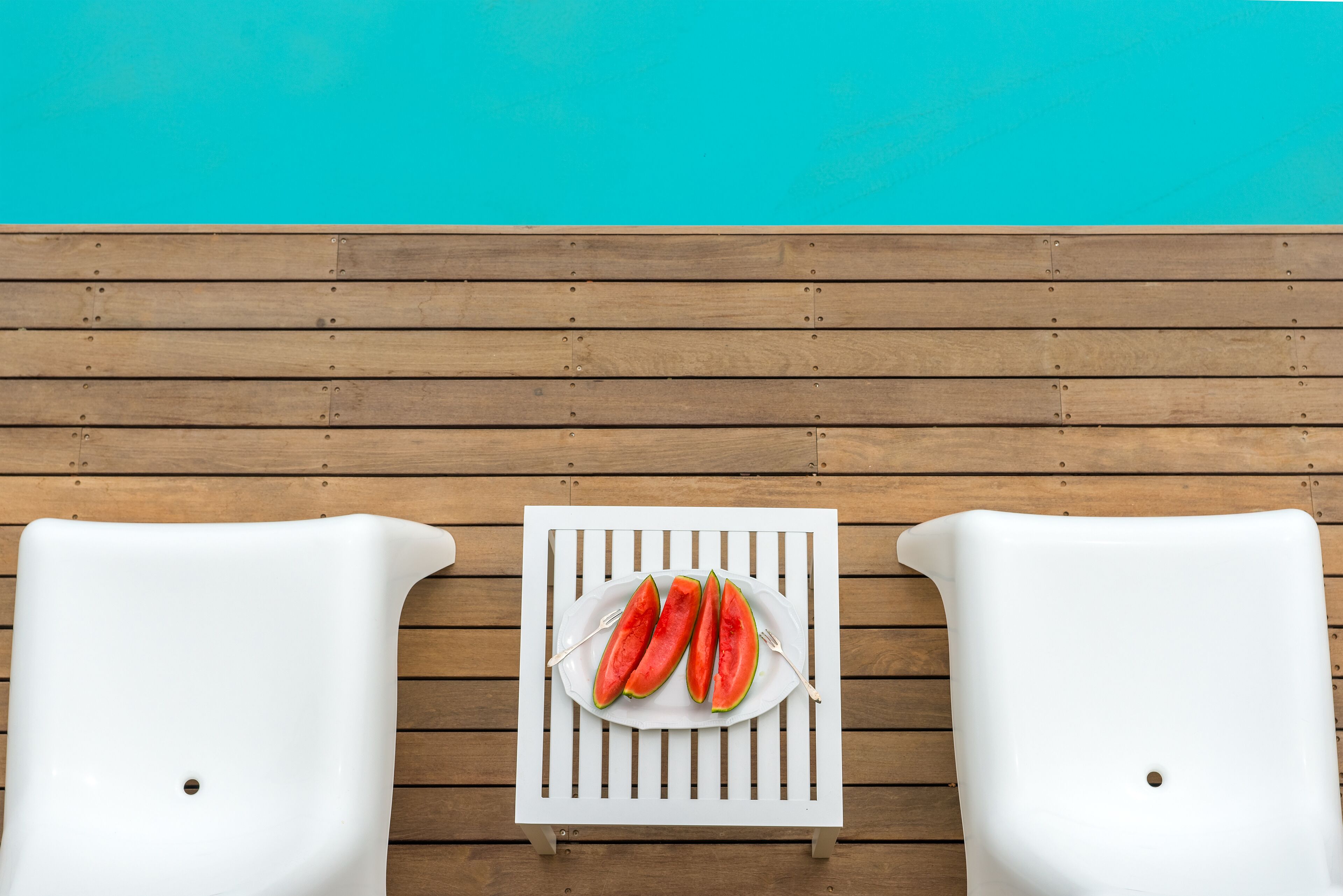 Área da piscina com duas espreguiçadeiras brancas e uma mesa branca com fatias de melancia.