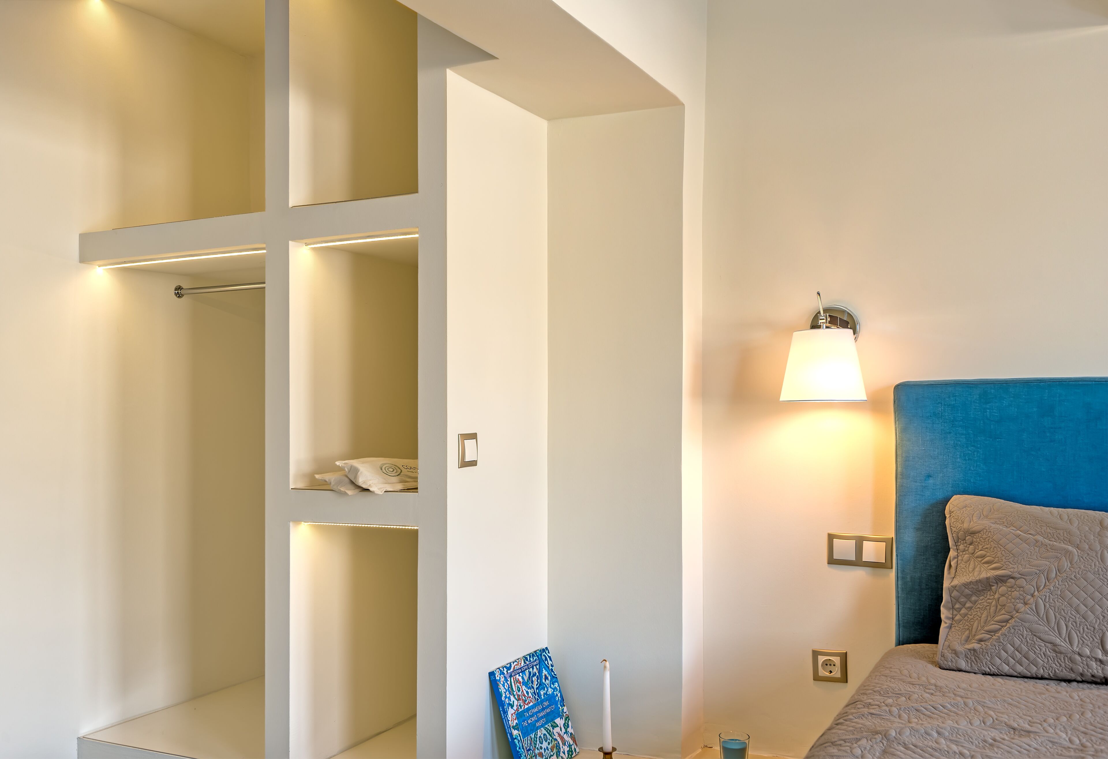 Quarto com cama, mesa de cabeceira, luminária na parede e estante com um livro.