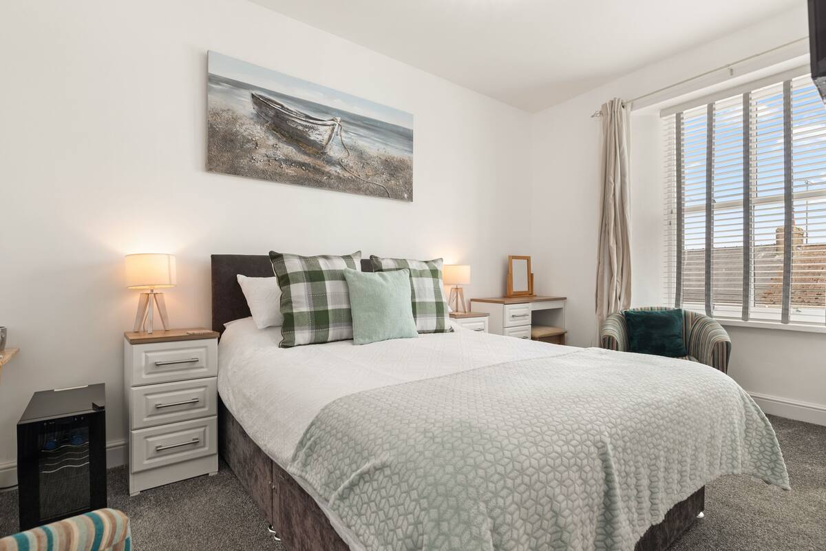 Quarto com cama, duas mesinhas de cabeceira, um criado-mudo, uma cadeira, janela com persianas e um quadro na parede.
