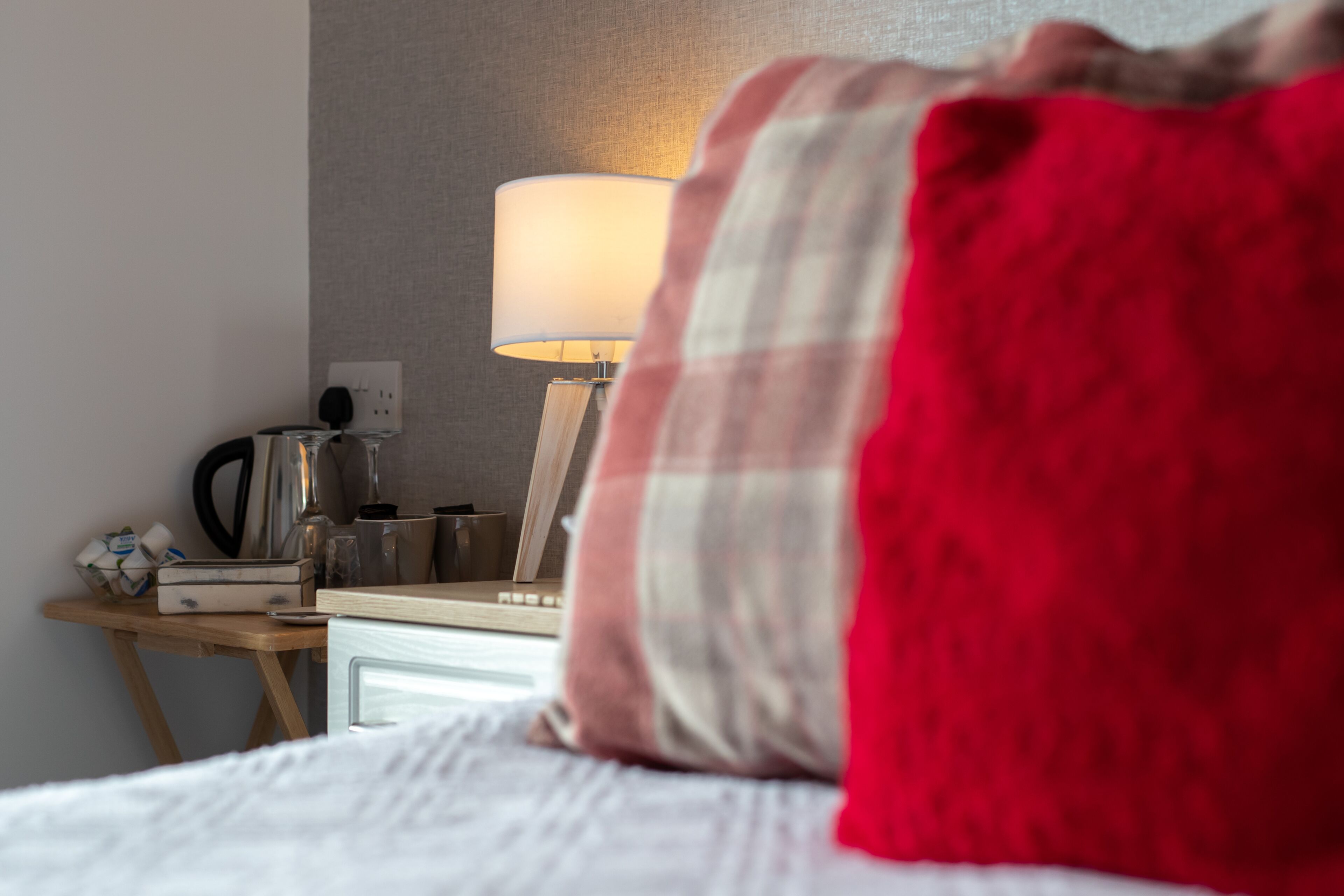 Cama bem arrumada com travesseiro vermelho e bege, mesa de cabeceira com abajur e pequeno criado-mudo com chaleira e outros objetos.