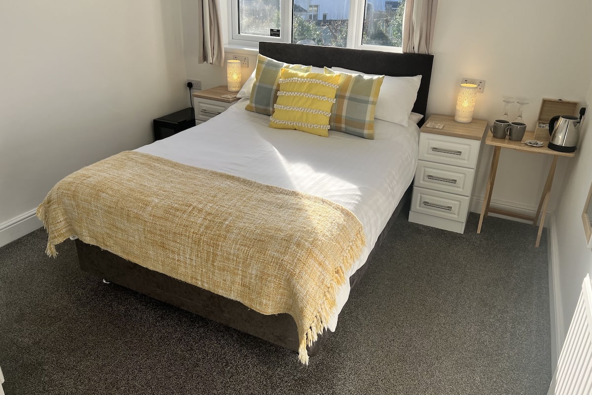 Cama bem arrumada com manta texturizada, luminária de cabeceira e mesa de apoio com chaleira e copos.