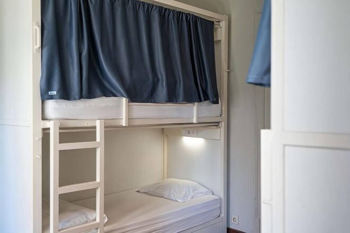 Uma beliche com escada, uma cama de solteiro embaixo e uma janela com cortinas.