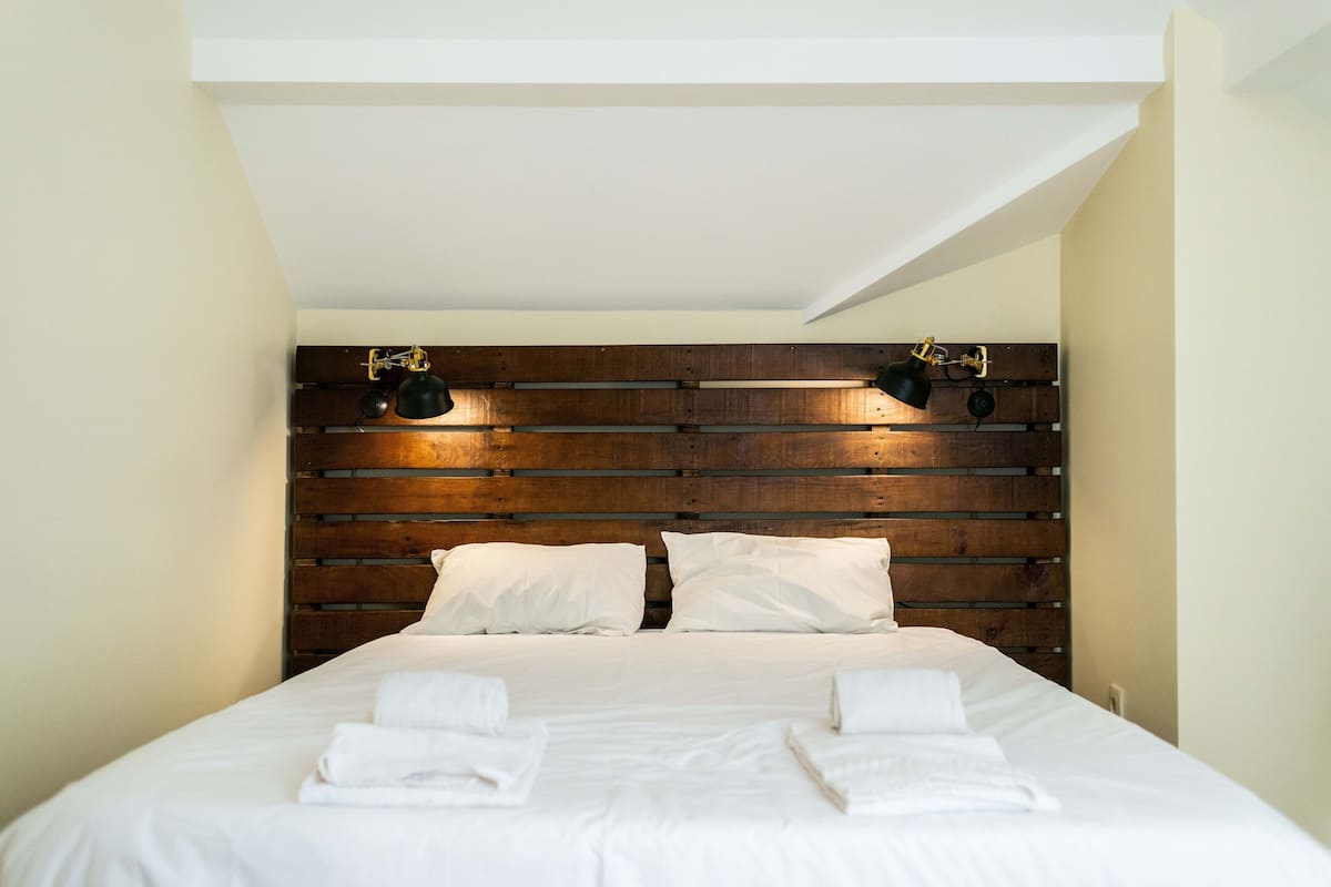 Cama bem arrumada com lençóis e toalhas brancos, cabeceira de madeira com ripas horizontais e duas luminárias fixadas na parede.