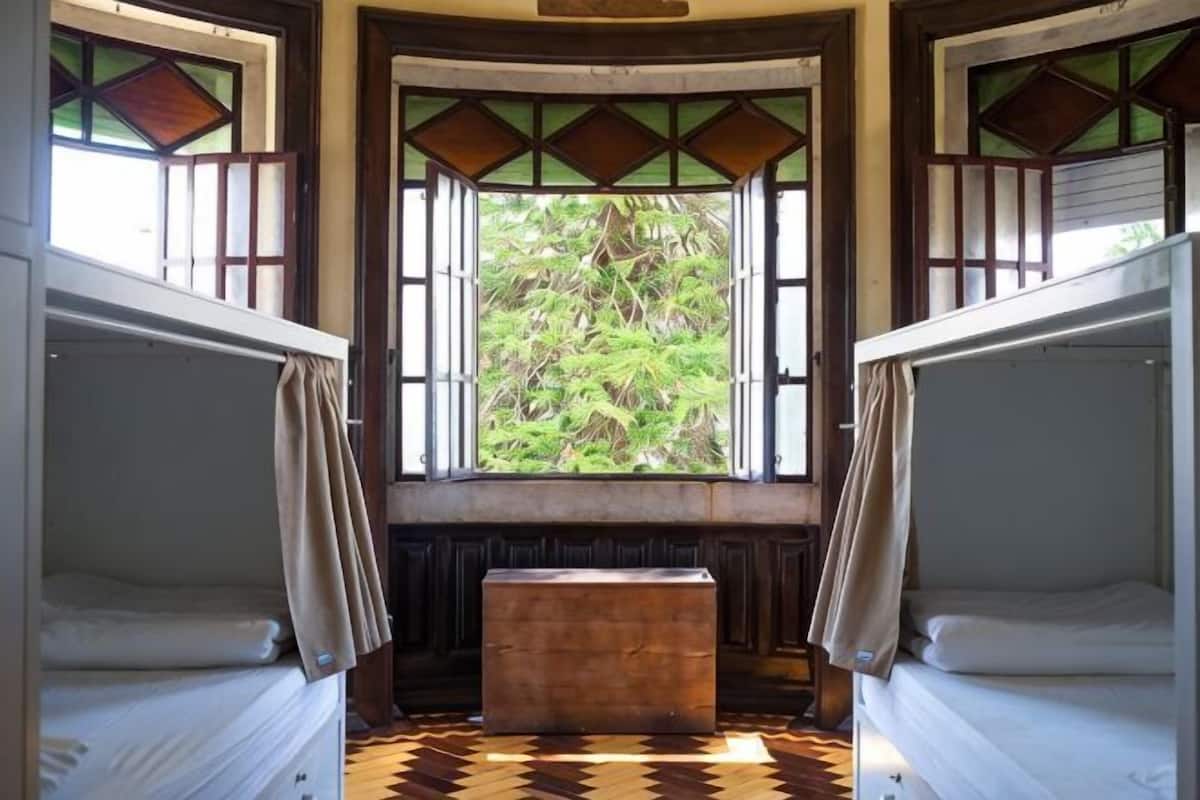 Quarto com duas camas de beliche, um banco e uma janela com vista para o verde.