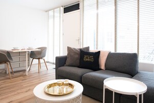 Uma sala de estar moderna com um sofá cinzento, uma mesa pequena branca e um espaço de jantar com uma mesa branca e duas cadeiras.