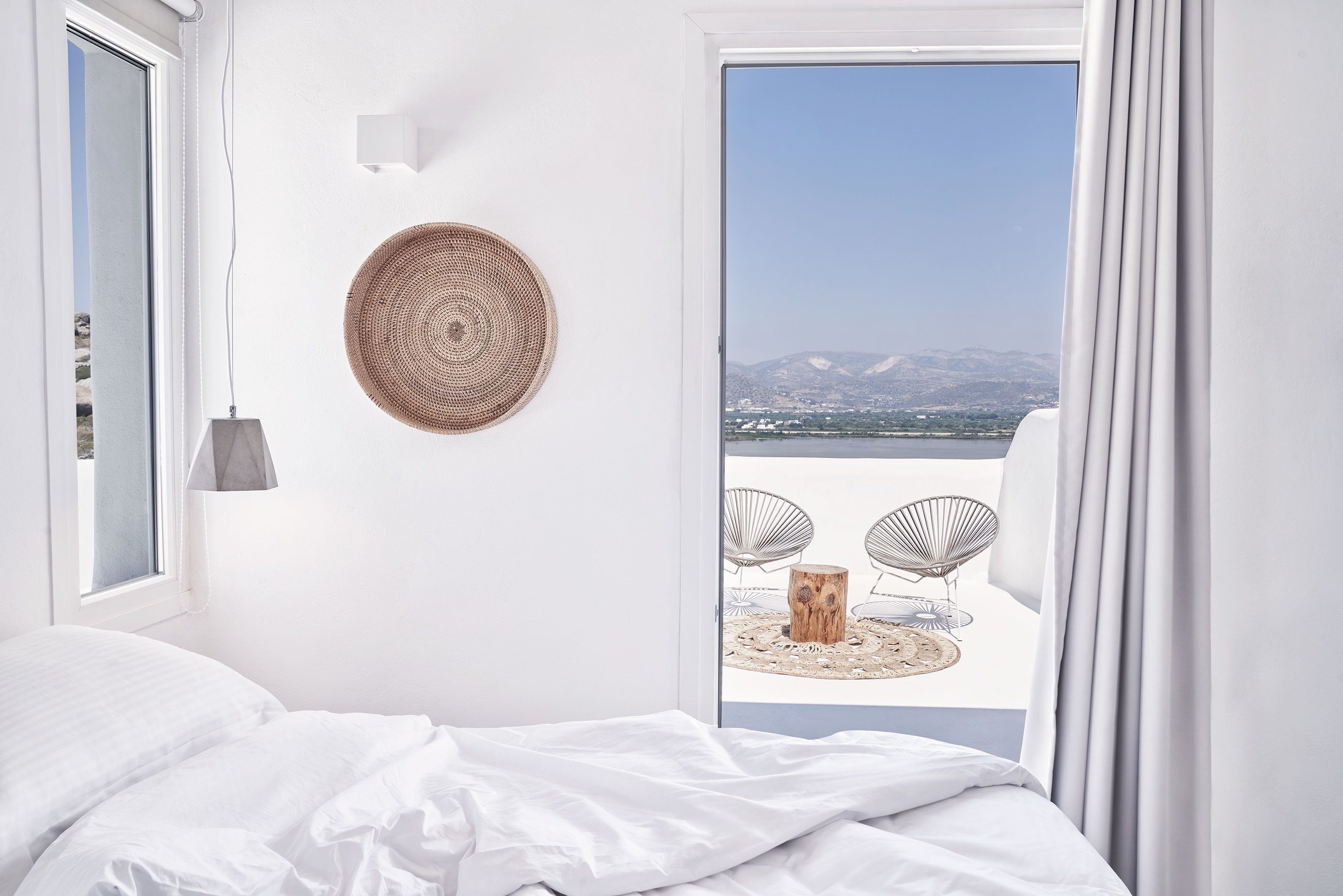 Quarto com cama, luminária na parede, arte em tear e vista para uma área de estar externa com cadeiras e mesa de madeira através de uma porta aberta.