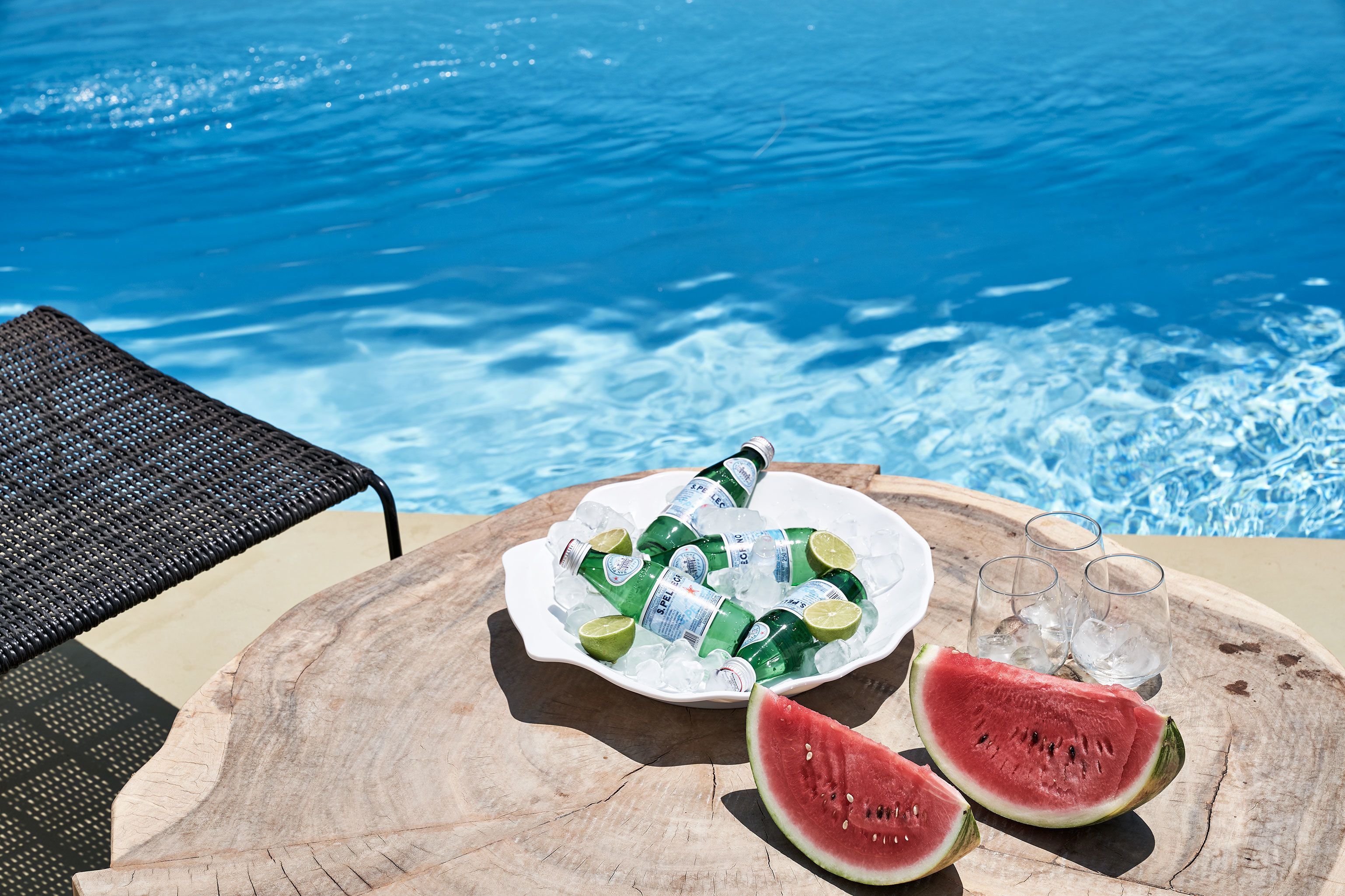 Uma mesa de madeira com uma travessa de melancias e garrafas, copos e uma cadeira perto de uma piscina.