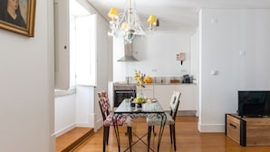 Uma área de refeições com mesa de vidro, cadeiras florais, um lustre e uma cozinha ao fundo.