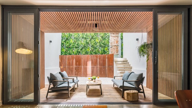 Um interior moderno com uma porta de vidro deslizante que dá para uma área externa com teto de madeira e parede verde. No interior, encontram-se duas cadeiras pretas, uma mesa de centro de madeira e um tapete pequeno.